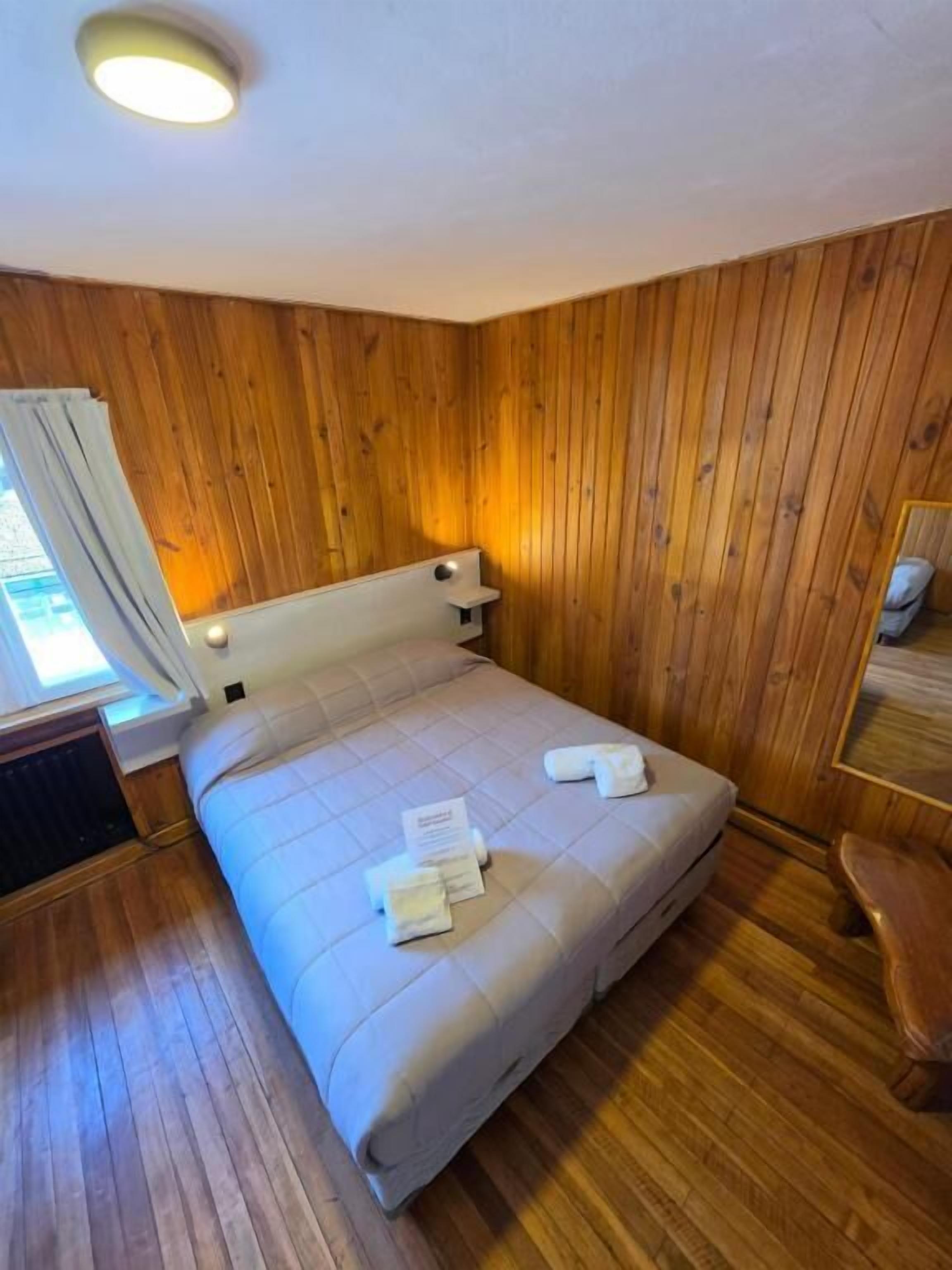 Quarto com painéis de madeira, uma cama com cabeceira, mesa de cabeceira e uma janela com cortinas.