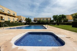 Duas piscinas sob um céu claro, um hedge e um edifício ao fundo.