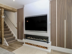 Uma sala de estar moderna com uma grande televisão de tela plana na parede branca, lareira com lenha, painéis de madeira nas paredes e uma escada com decoração de estrela-do-mar.