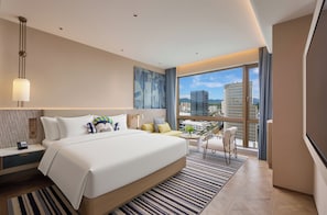 Quarto de hotel moderno com uma cama grande, uma escrivaninha, uma cadeira, uma mesa pequena e vista para a cidade através de grandes janelas.