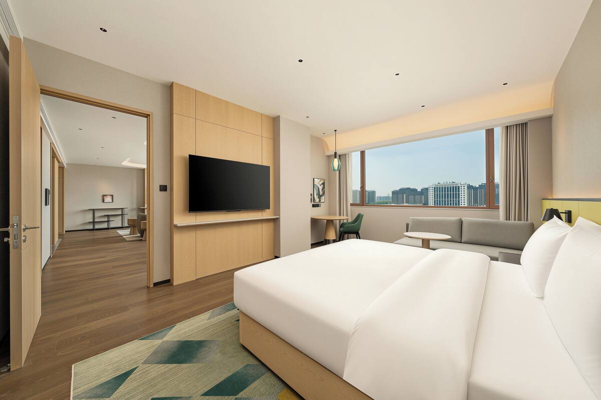 Quarto de hotel com uma cama grande, televisão na parede, sofá, mesinha e vista para a cidade.