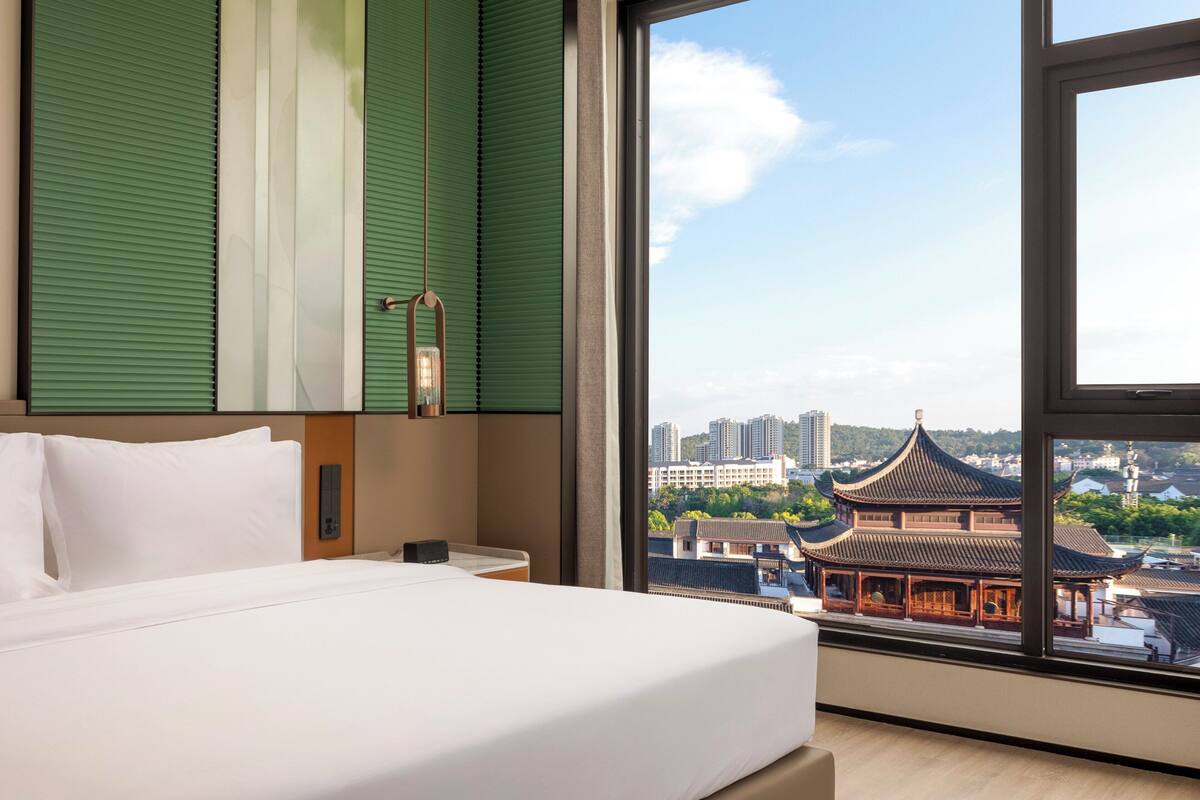 Quarto de hotel com cama, mesa de cabeceira, luminária e uma grande janela com vista para arquitetura tradicional.