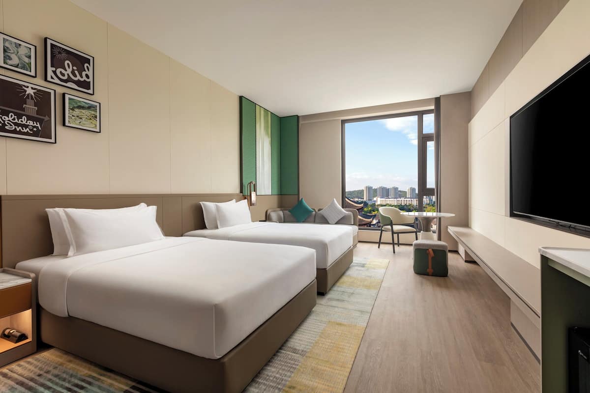 Quarto de hotel com duas camas, televisão, varanda com vista para a cidade e quadros nas paredes.