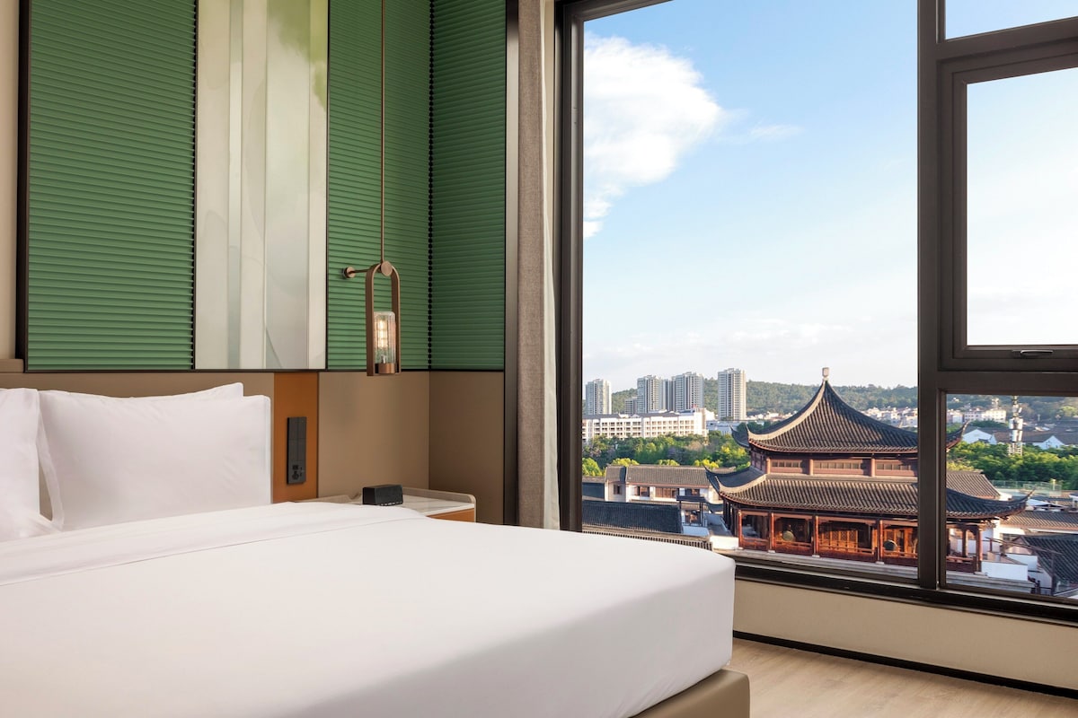 Quarto de hotel com cama, mesa de cabeceira, luminária e uma grande janela com vista para arquitetura tradicional.