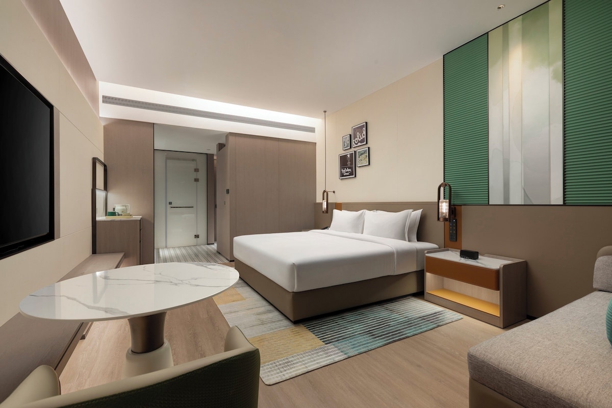 Quarto de hotel moderno com cama, escrivaninha, cadeira, televisão e uma janela grande.
