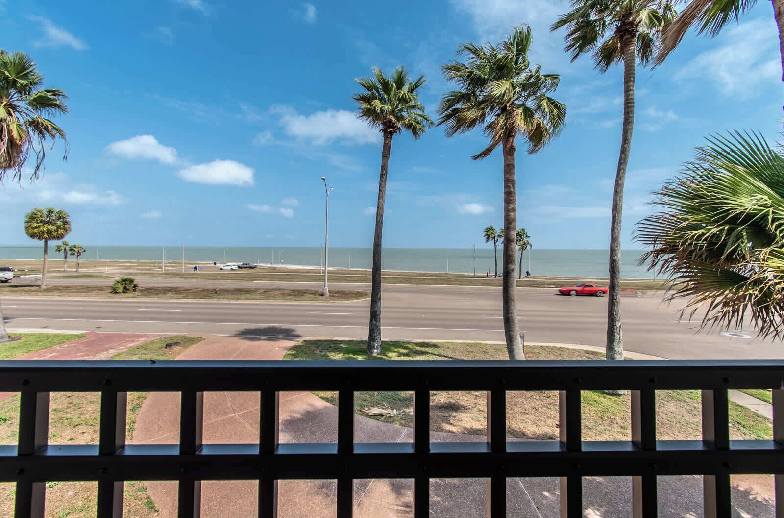 Vista de uma varanda com guarda-corpo, com uma estrada, palmeiras e uma praia ao longe.