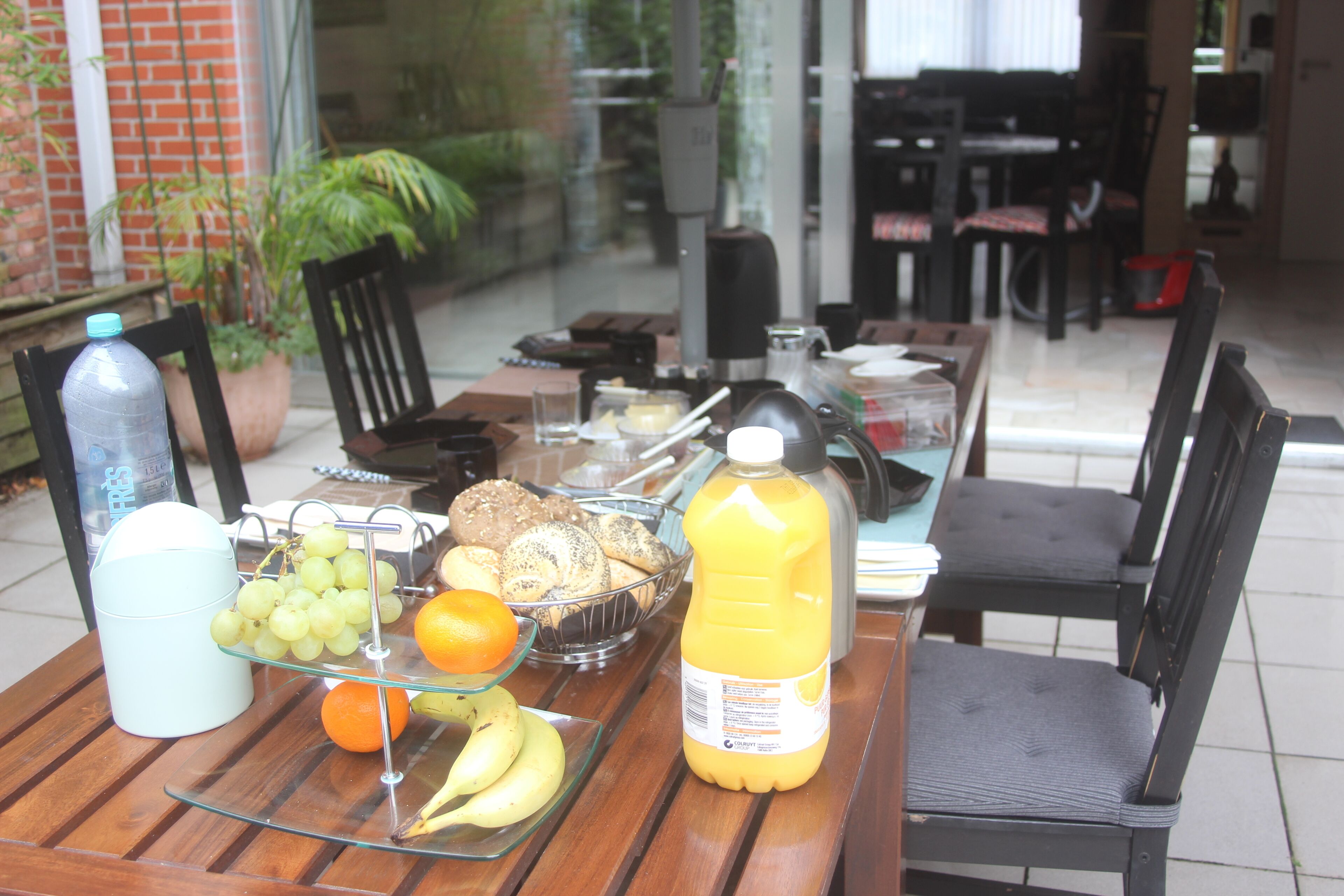 Uma mesa de jantar com uma tigela de vidro cheia de uvas, uma laranja e bananas, uma garrafa de suco de laranja e uma garrafa de água.