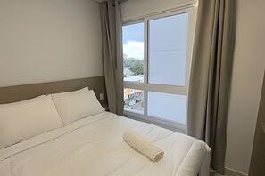 Quarto de hotel com uma janela grande, uma cama bem arrumada e vista para a paisagem urbana.