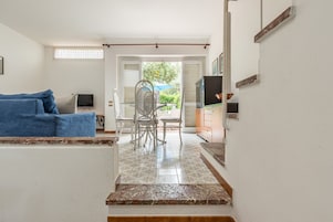 Um corredor que leva a uma sala de jantar com mesa e cadeiras, um sofá azul, uma televisão e uma escada.