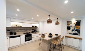 Uma cozinha moderna com armários brancos, uma ilha central com banquetas, máquina de café e luminárias pendentes.