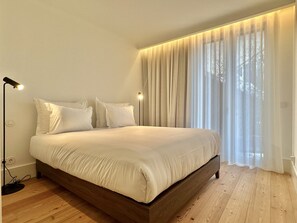 Quarto com cama de madeira, roupa de cama branca, abajur e cortinas translúcidas.