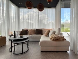 Uma sala de estar moderna com um sofá seccional, uma mesa de centro redonda e vista para um jardim através de grandes janelas.