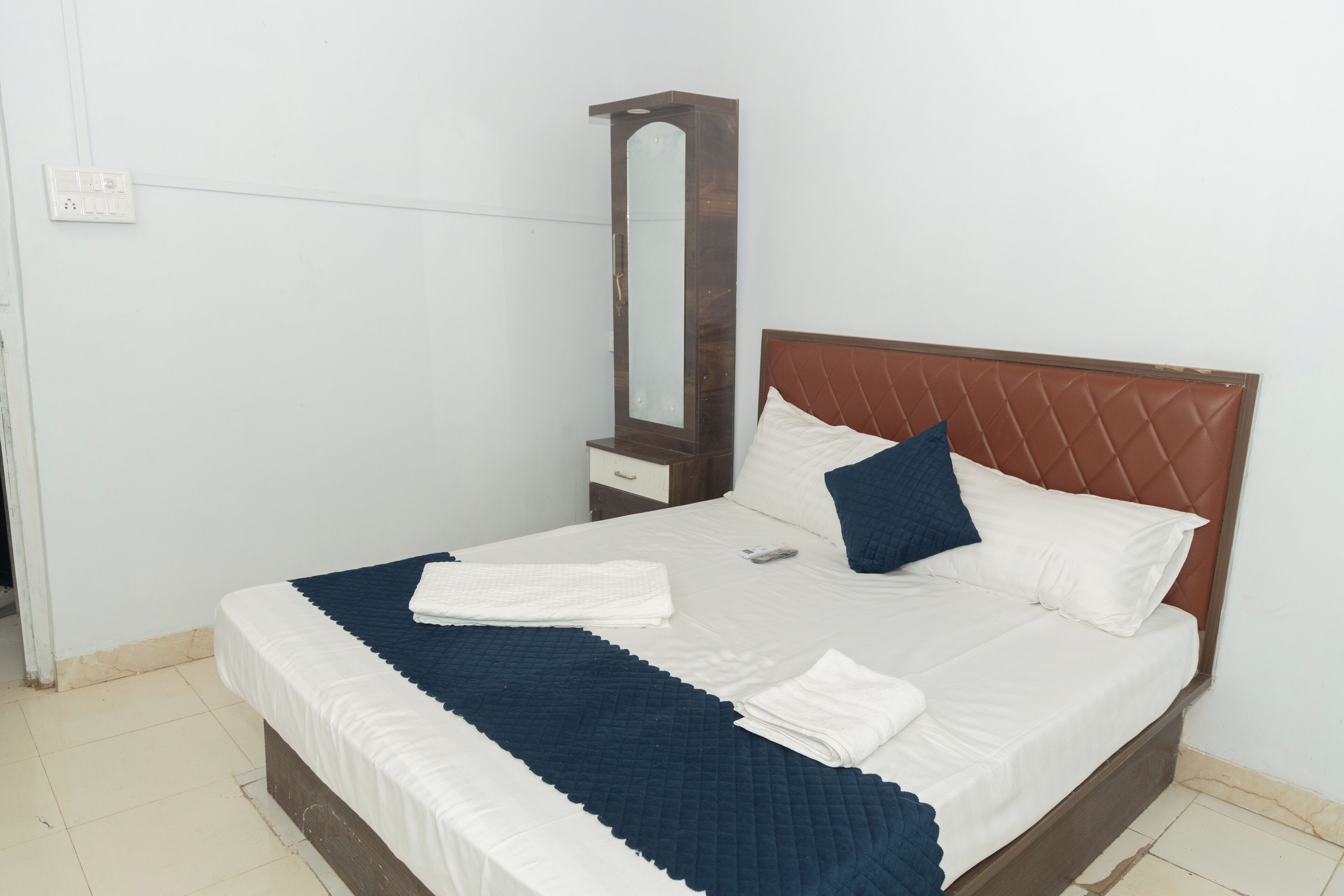 Cama bem arrumada com roupa de cama branca e azul, cabeceira de madeira, espelho de cabeceira e uma pequena gaveta.