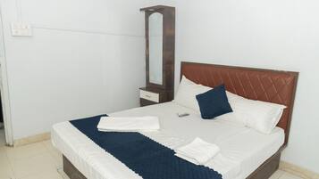 Cama bem arrumada com roupa de cama branca e azul, cabeceira de madeira, espelho de cabeceira e uma pequena gaveta.