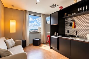 Quarto de hotel moderno com sofá, área de jantar com bancada de cozinha, janela com vista para a cidade e luminária pendente.