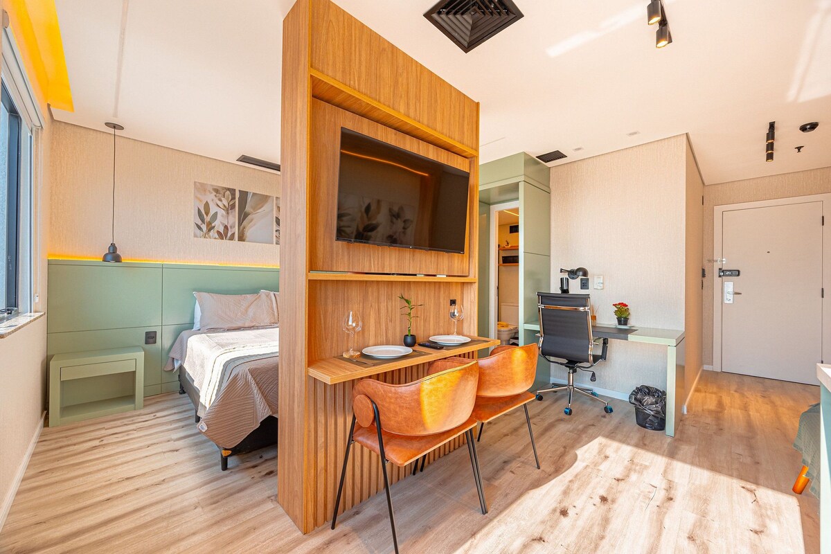 Quarto de hotel compacto com uma cama, uma escrivaninha de madeira, duas cadeiras, uma televisão e uma pequena mesa com uma planta.
