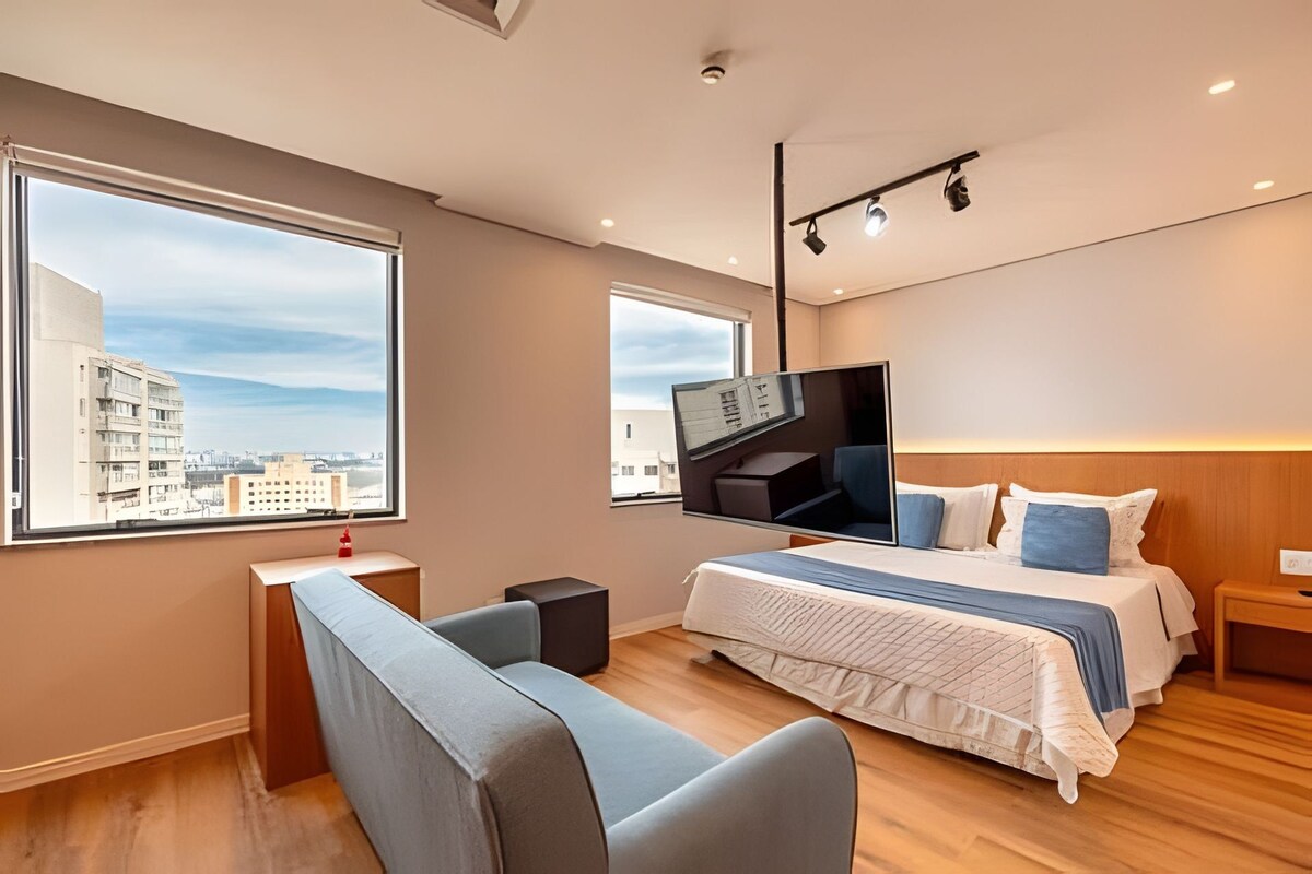 Quarto de hotel moderno com uma cama grande, área de estar, televisão na parede e vista para a cidade pelas janelas.