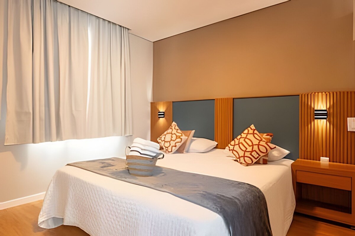 Quarto de hotel com cama, mesas de cabeceira e janela com cortinas.