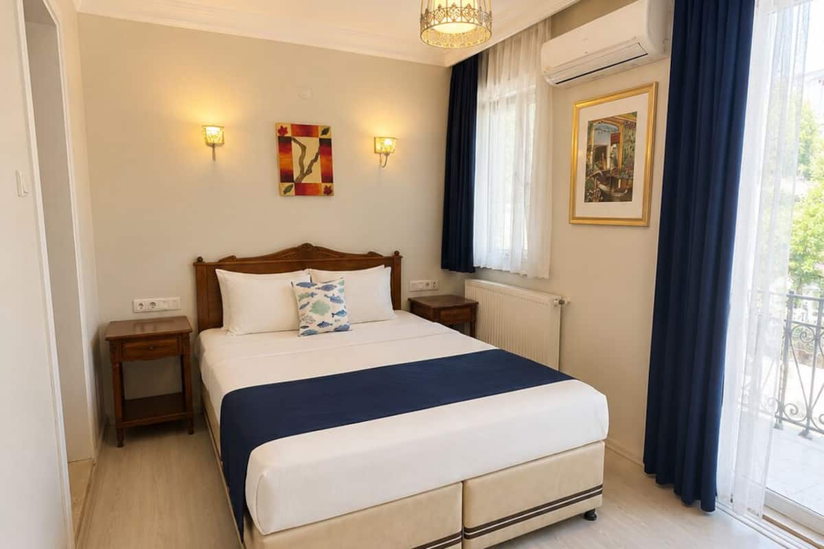Quarto de hotel com cama, mesas de cabeceira, um banquinho vermelho, luminária no teto e janela com cortinas.