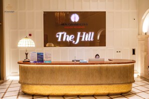 Balcão de recepção com uma TV de tela plana exibindo o letreiro 'The Hill', uma luminária e folhetos informativos.