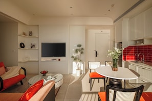 Uma sala moderna com televisão de tela plana, área de jantar com mesa redonda e cadeiras, e cozinha com armários brancos e azulejos vermelhos.