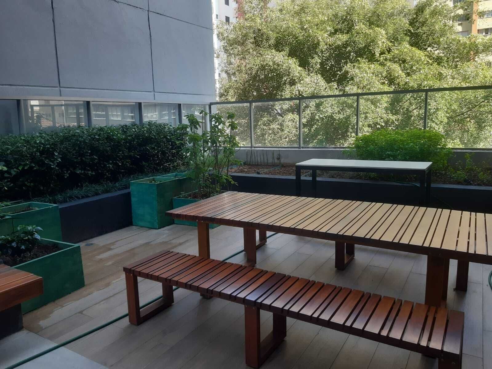 Um pátio externo com mesa e bancos de madeira, cercado por vegetação e vista para árvores.