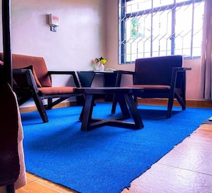 Quarto com carpete azul, dois sofás marrons, uma mesinha com um vaso e uma janela com grades.