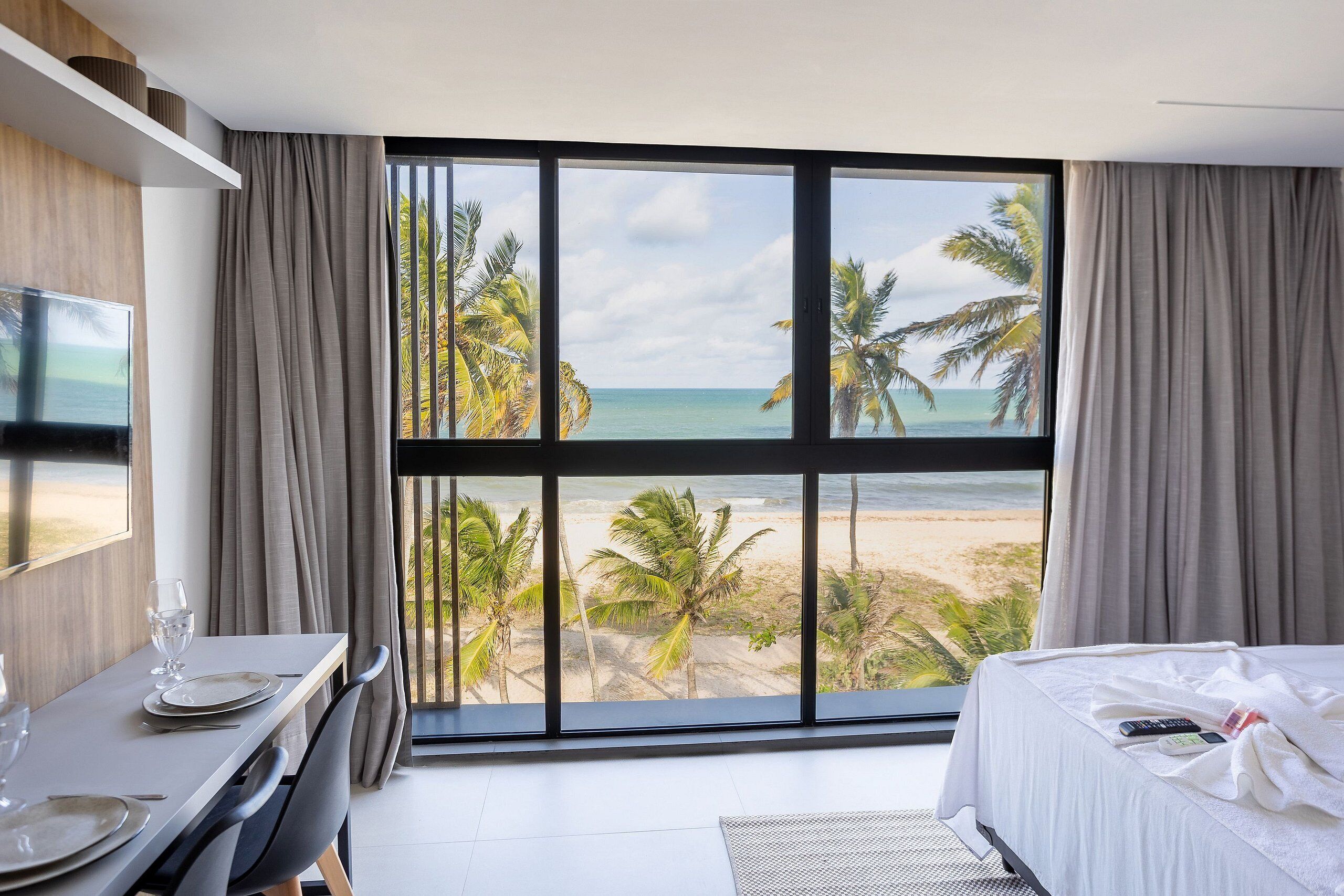 Quarto de hotel com uma janela grande, uma cama, uma escrivaninha com cadeira e uma televisão.