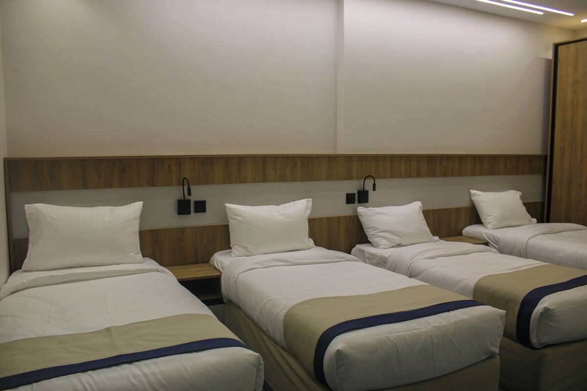 Quarto de hotel com quatro camas de solteiro, cada uma com cabeceira de madeira, roupa de cama branca e um toque de azul no edredom.