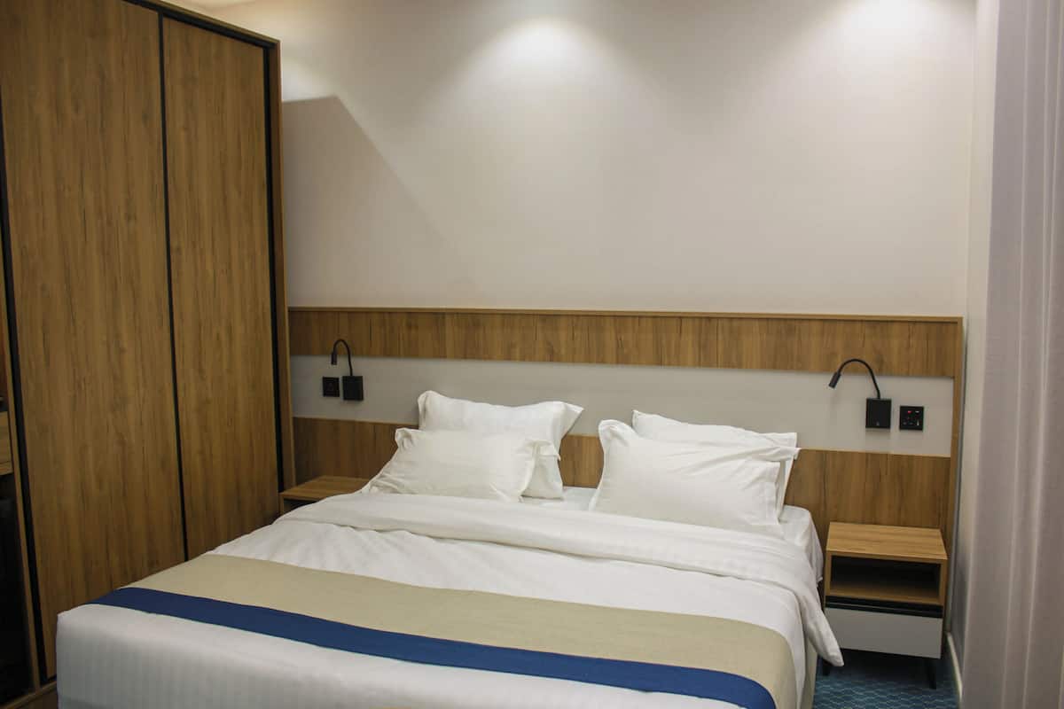 Quarto de hotel com cama, guarda-roupa de madeira, mesa de cabeceira e luminária na parede.