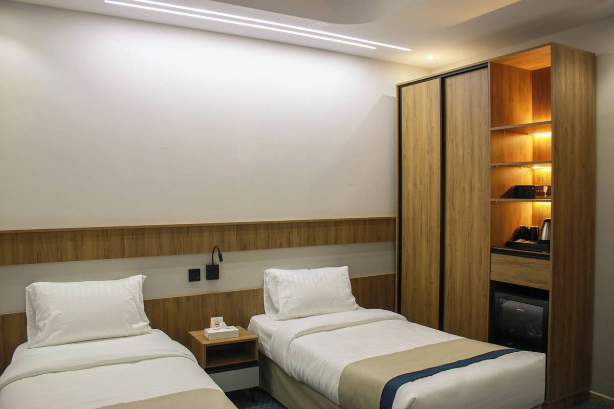 Quarto de hotel com duas camas, um guarda-roupa de madeira, uma escrivaninha com telefone e uma pequena prateleira com objetos.