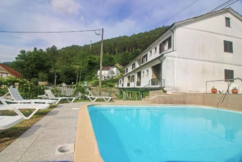 Uma piscina com espreguiçadeiras, um edifício com várias janelas e uma encosta arborizada ao fundo.