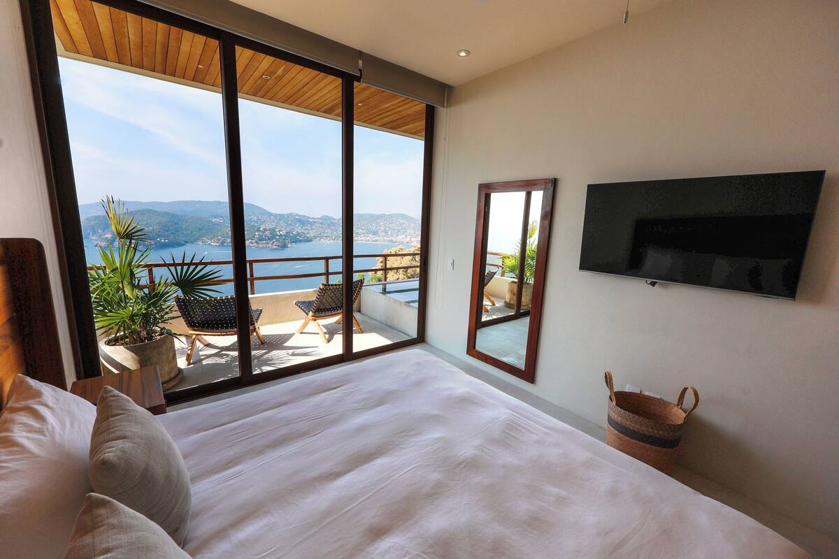 Quarto com cama grande, televisão na parede e varanda com vista para o mar.