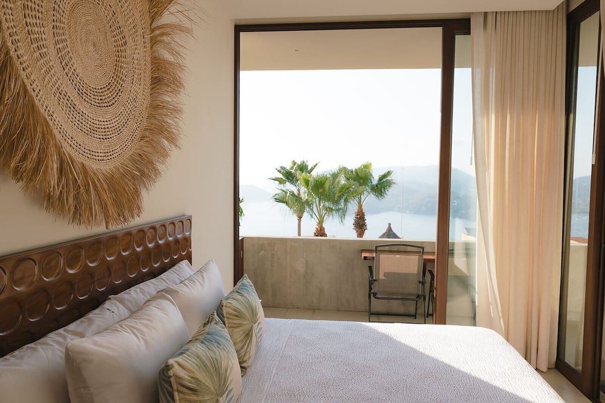 Quarto com uma cama grande, vista para palmeiras e para a água, e varanda com uma cadeira.