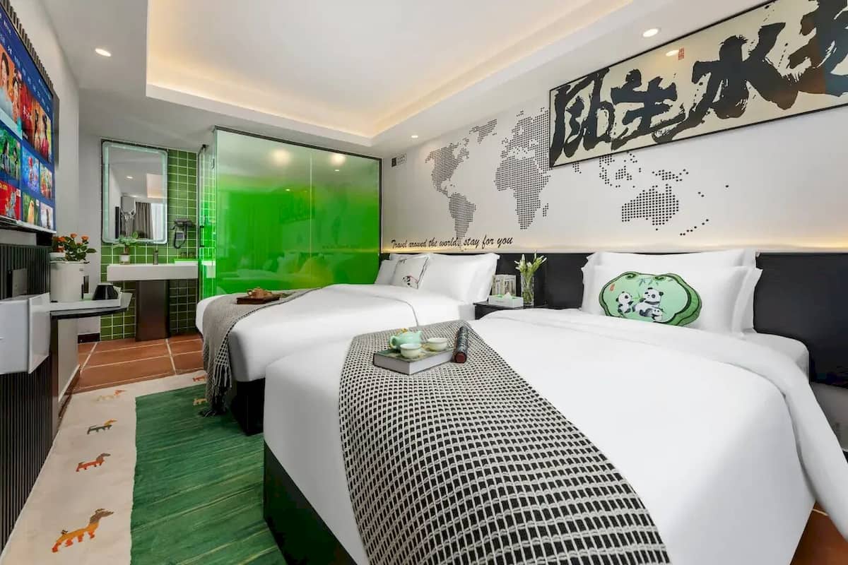 Quarto de hotel moderno com duas camas, uma parede de destaque verde, um mapa-múndi e uma pequena área de trabalho.