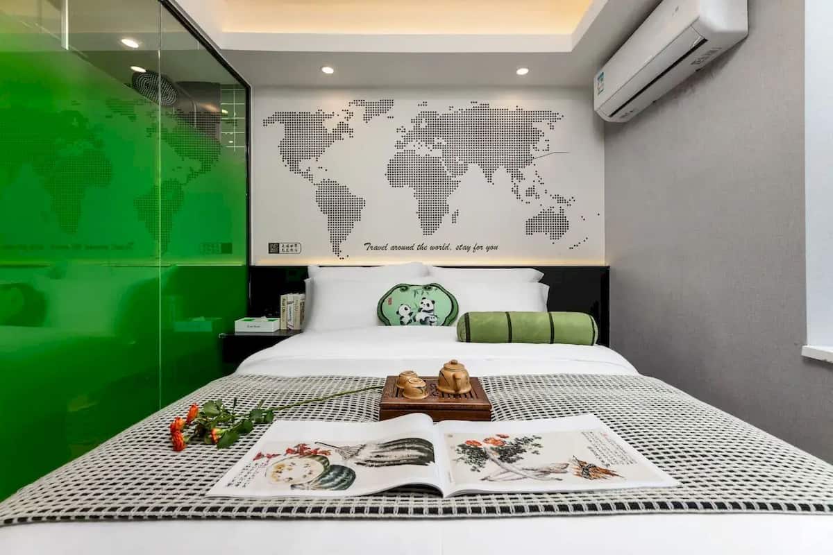 Quarto de hotel moderno com um mural de mapa-múndi, uma cama com almofada verde e uma revista sobre a cama.