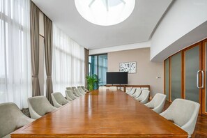 Uma sala de conferências com uma longa mesa de madeira, cadeiras bege, uma janela grande com cortinas, uma televisão e um mapa na parede.
