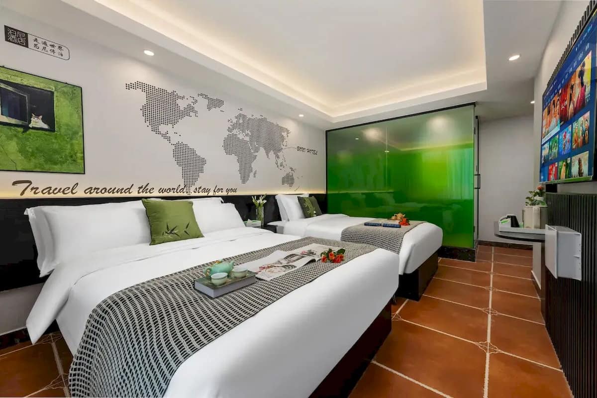 Quarto de hotel com duas camas, um mapa-múndi na parede, uma parede de destaque verde e um balcão de recepção com uma tela de televisão.