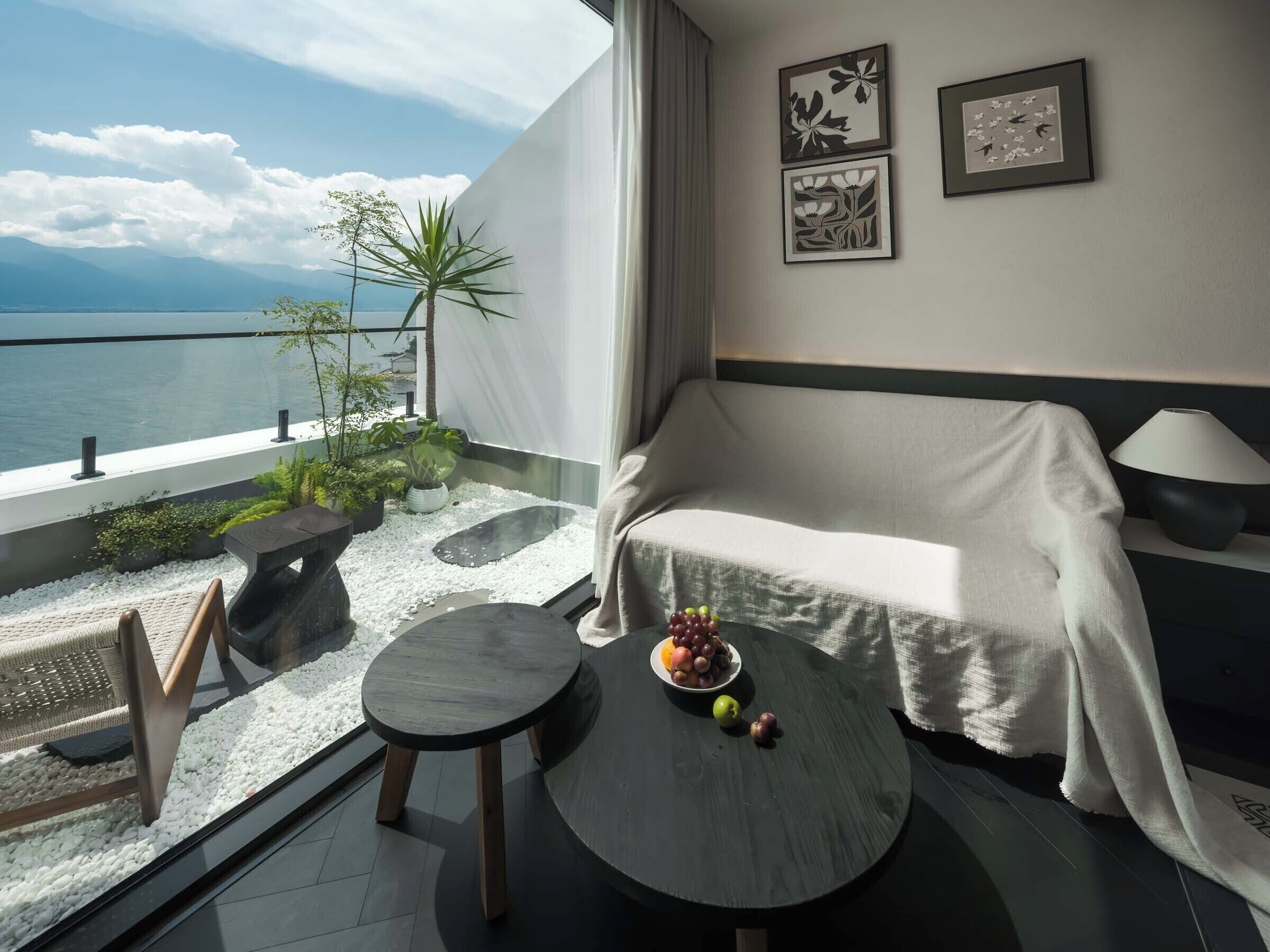 Varanda com vista para o mar, um sofá, uma mesa redonda pequena com frutas e duas obras de arte emolduradas na parede.