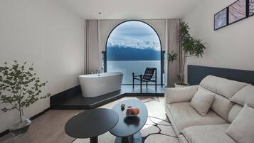 Uma sala de estar moderna com sofá, mesa de centro redonda, banheira e vista para o mar por uma janela em arco.