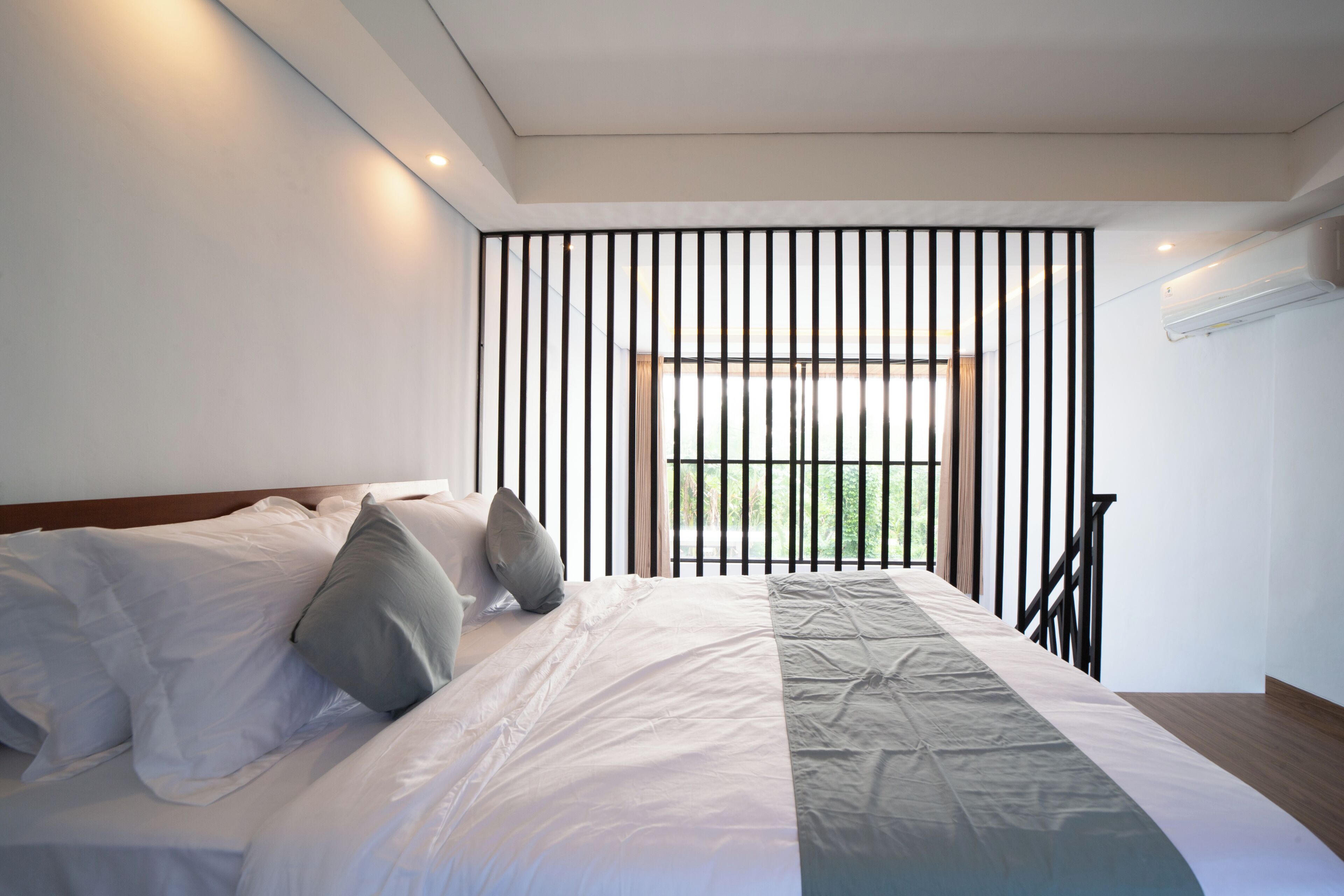 Quarto moderno com uma cama grande, vista pela janela com persianas verticais e uma escada que leva a um andar superior.