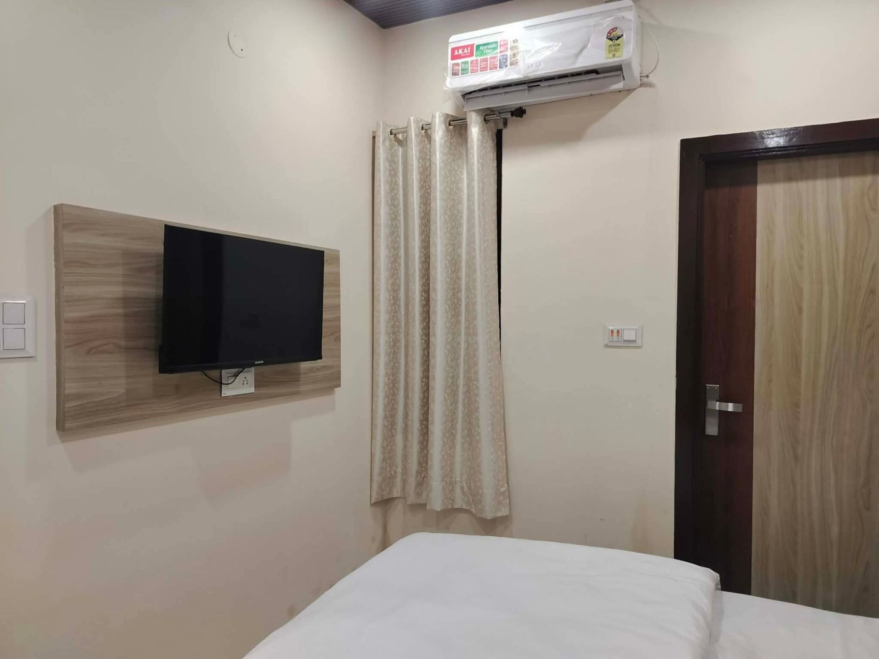 Quarto de hotel com televisão montada, ar condicionado, cortina e porta de madeira.