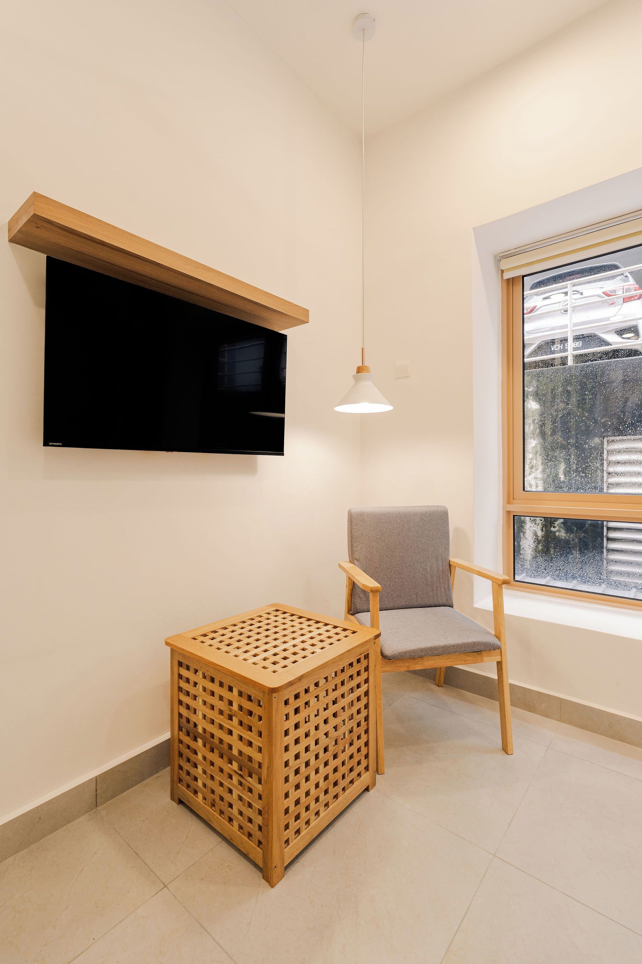 Quarto com televisão na parede, uma cadeira de madeira, uma mesa pequena de madeira e janela com vista para carros estacionados.