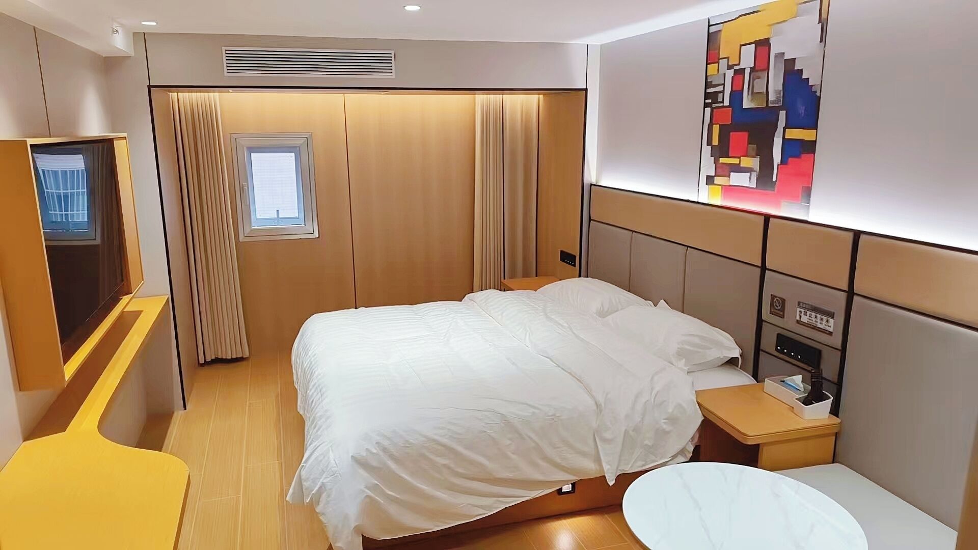 Quarto de hotel com cama, mesa de cabeceira, poltrona, janela com cortinas, obra de arte na parede e ar condicionado embutido.