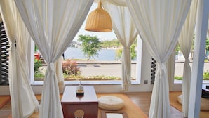 Quarto com vista para um corpo d'água, cortinas brancas, luminária pendente trançada, mesa lateral de madeira e um banquinho otomano trançado.