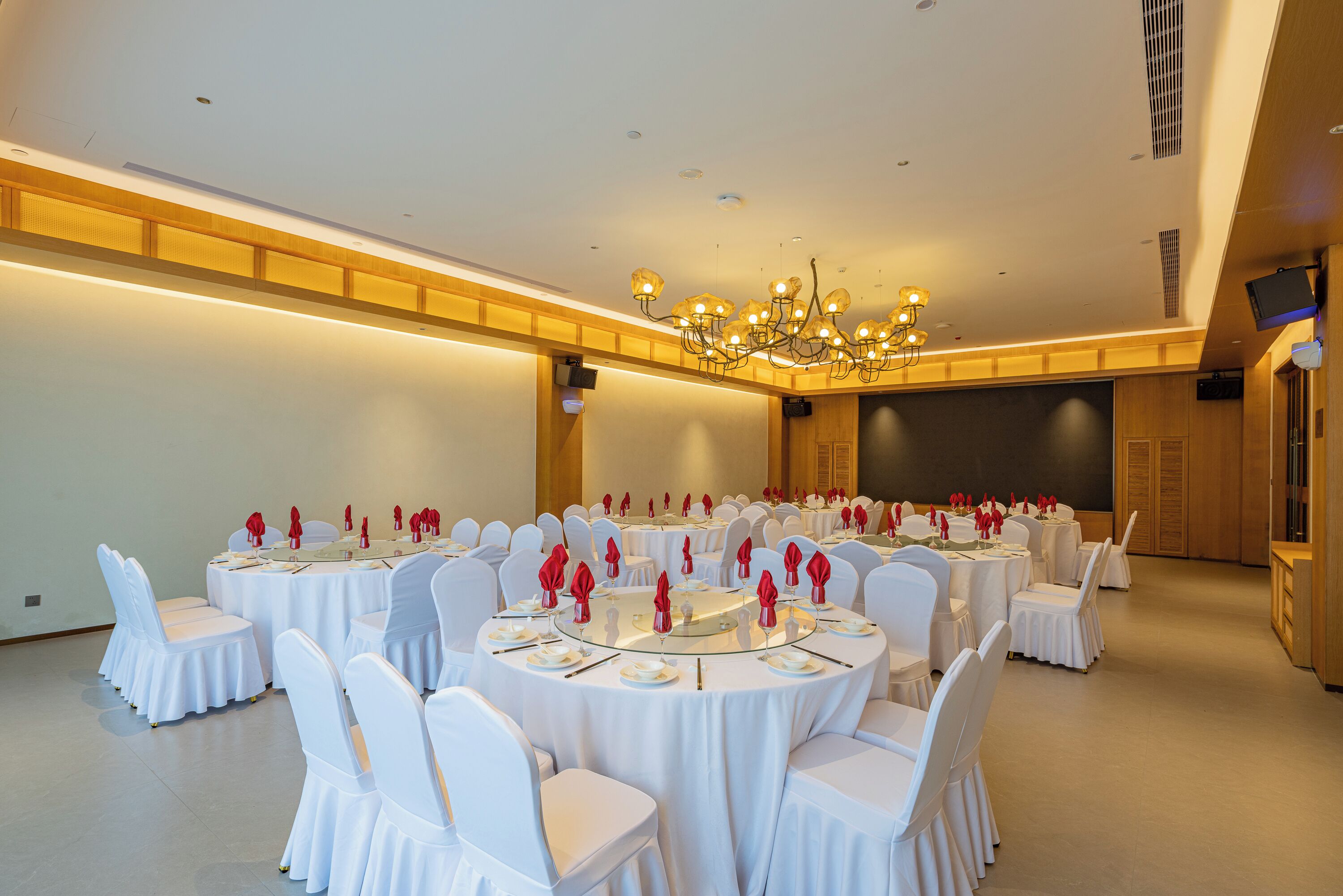 Uma sala de banquetes com várias mesas redondas, cadeiras brancas e guardanapos vermelhos.