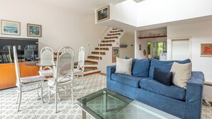 Uma sala de estar com um sofá azul, cadeiras brancas, uma mesa de centro de vidro, uma escada e um espaço de jantar com uma mesa e cadeiras de madeira.