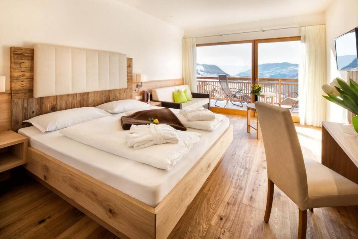 Quarto de hotel com cama de madeira, varanda com vista, escrivaninha e uma cadeira.