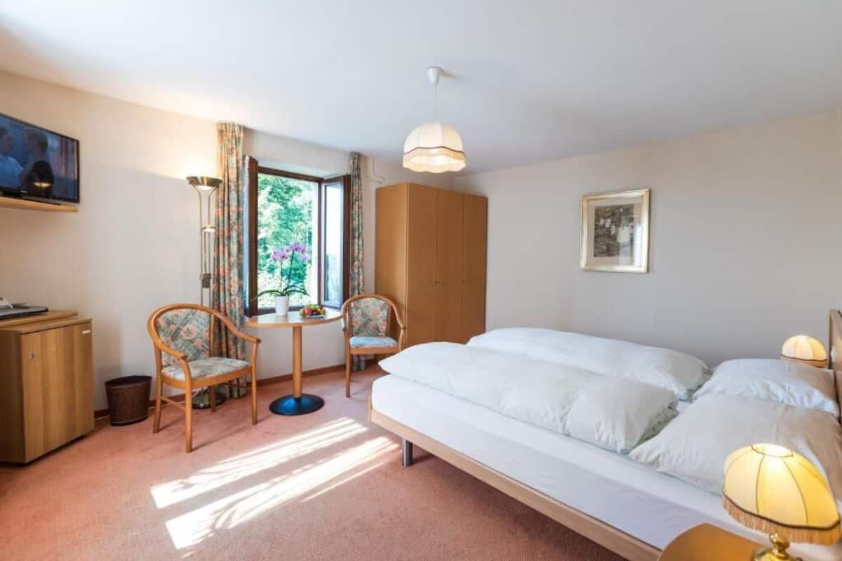 Quarto com cama, duas cadeiras, uma mesinha, guarda-roupa, televisão e janela com cortinas.