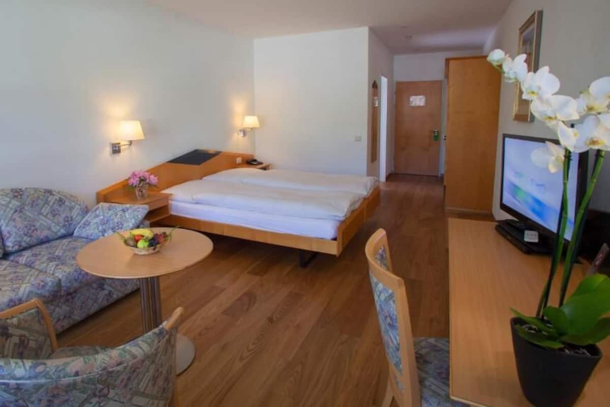 Quarto com cama, sofá, mesinha com uma tigela de frutas, cadeira, televisão e planta em vaso.